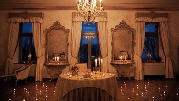 Uma sala de jantar formal com candelabro, espelhos ornamentados e velas no chão.