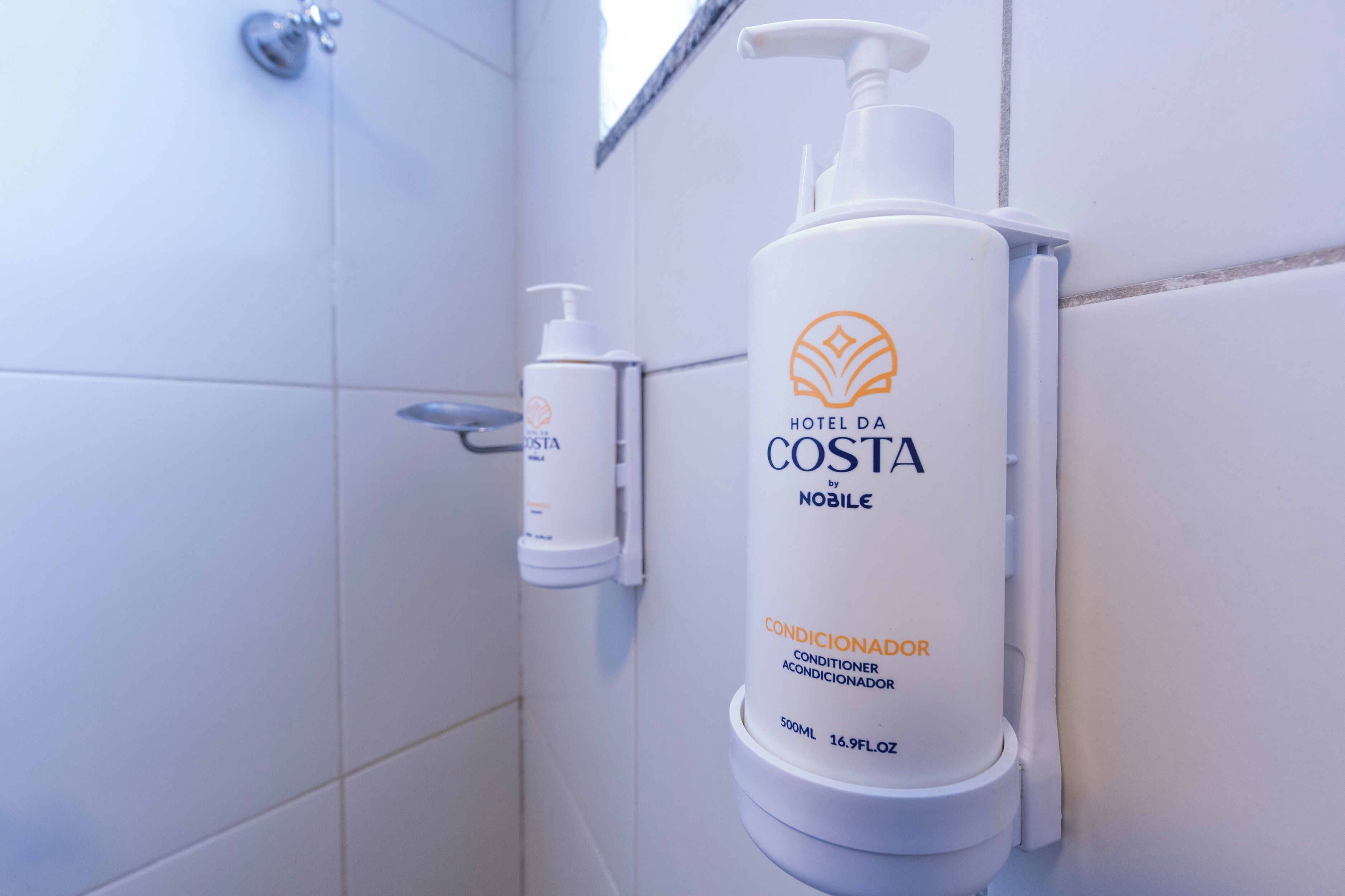 Um banheiro com azulejos brancos, chuveiro e dois frascos de condicionador Hotel da Costa by Noble.