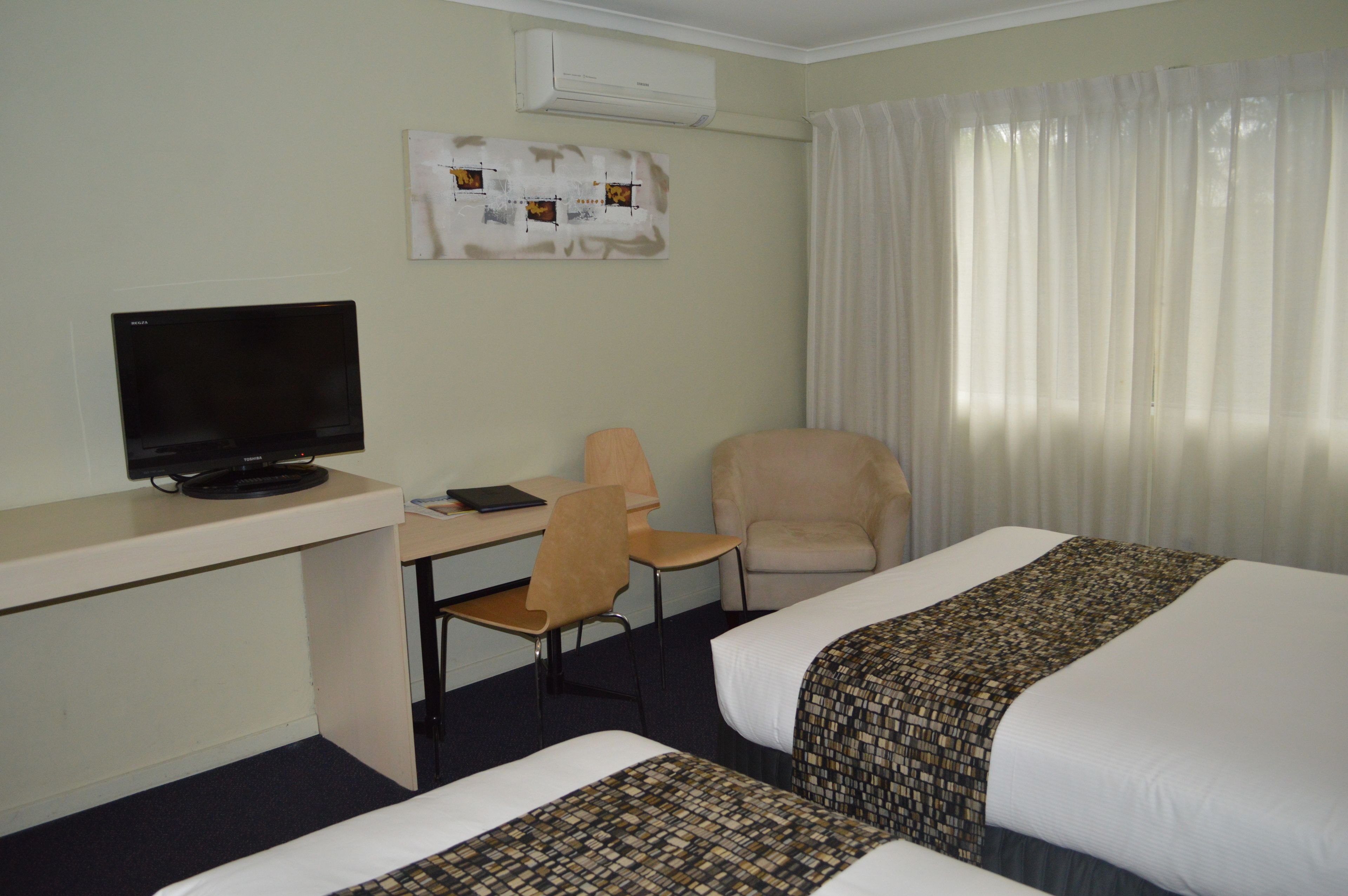 Quarto de hotel com escrivaninha, televisão, duas camas, uma cadeira e janela com cortinas.
