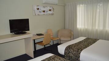 Quarto de hotel com escrivaninha, televisão, duas camas, uma cadeira e janela com cortinas.