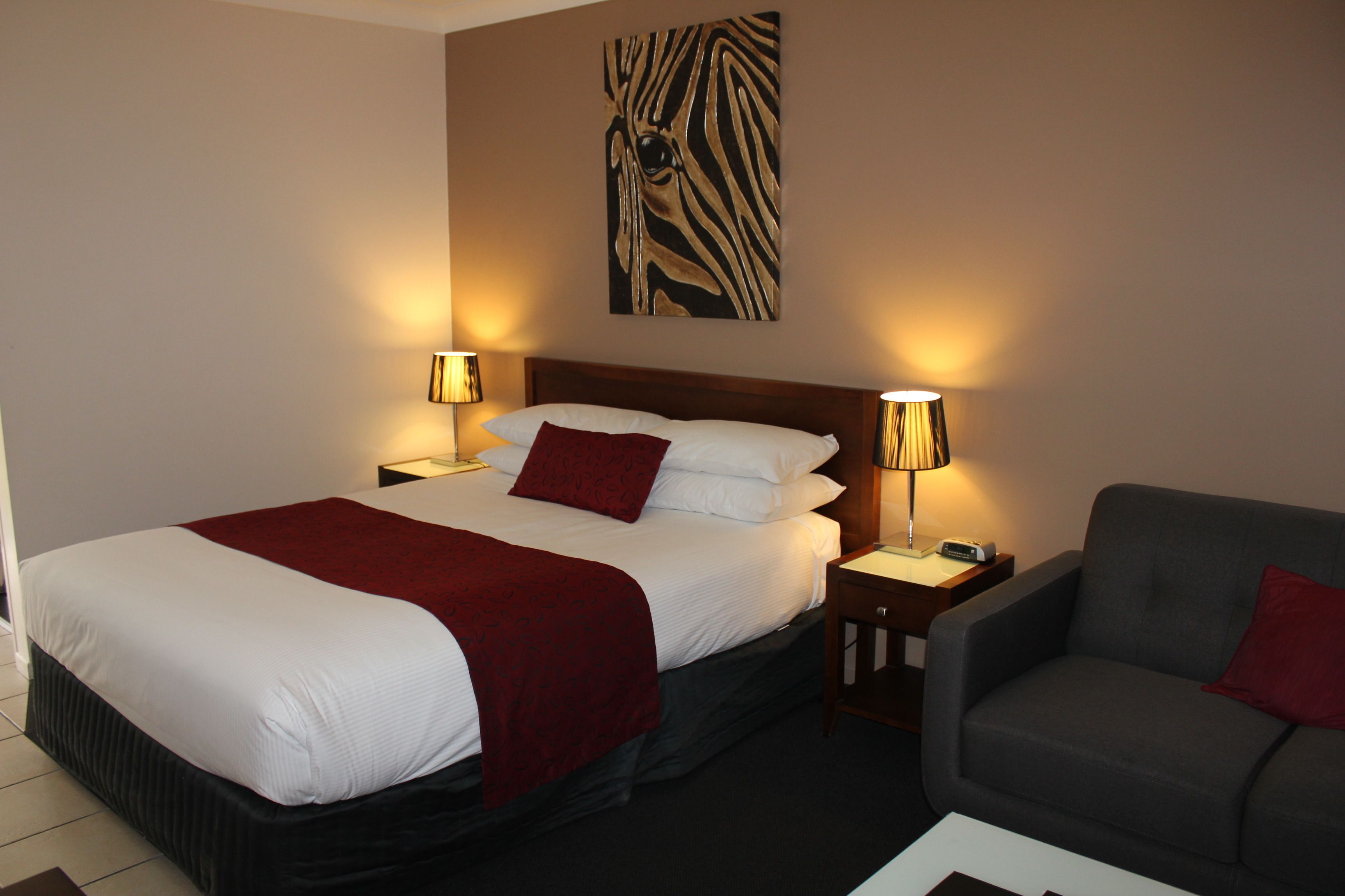 Quarto de hotel com uma cama grande, duas mesas de cabeceira com luminárias, uma poltrona cinza e uma obra de arte na parede com padrão de zebra.
