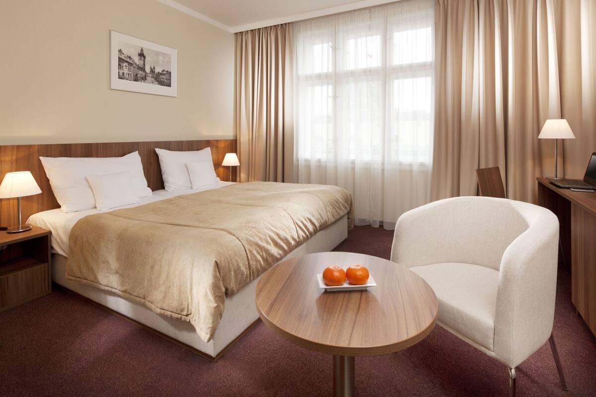 Quarto de hotel com cama, duas luminárias de cabeceira, uma mesa redonda com laranjas, uma poltrona branca e um laptop sobre uma escrivaninha.