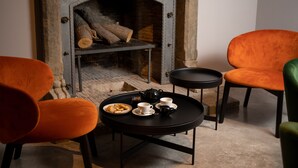 Um quarto aconchegante com lareira, duas poltronas laranja, uma mesa pequena com uma bandeja de comida, xícaras e um bule.