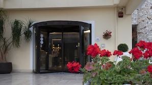 Entrada de hotel com porta giratória, plantas em vasos e flores vermelhas.