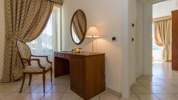 Quarto de hotel com escrivaninha de madeira, cadeira, luminária, espelho e vista para o exterior através de uma janela com cortinas.