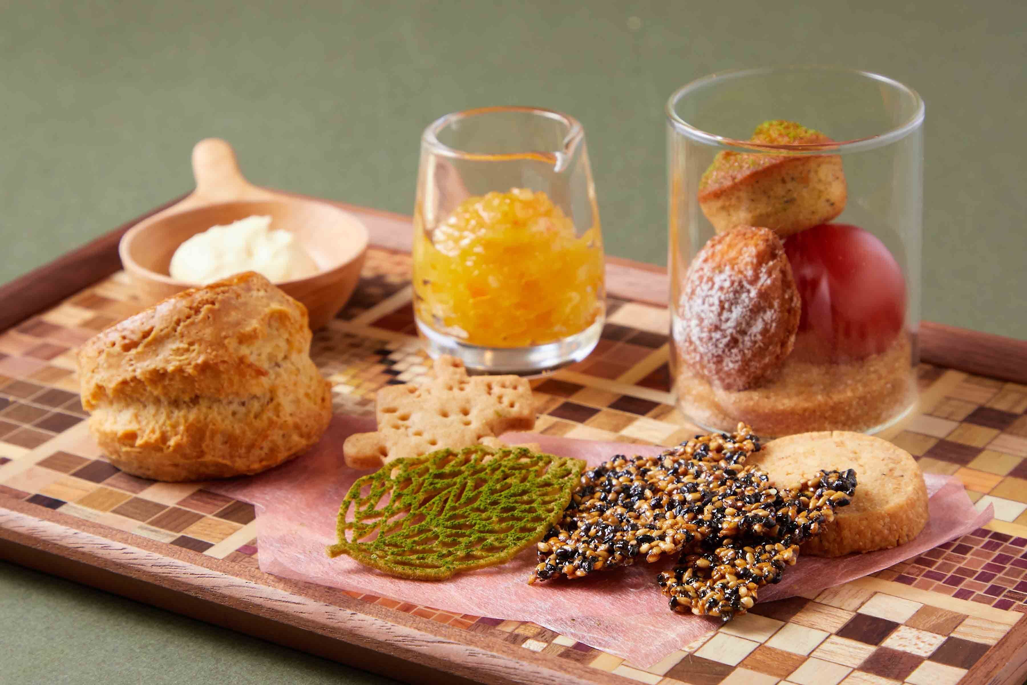 Uma bandeja com diversos produtos de panificação, incluindo scones, um doce em formato de folha verde e um copo com um líquido amarelo.