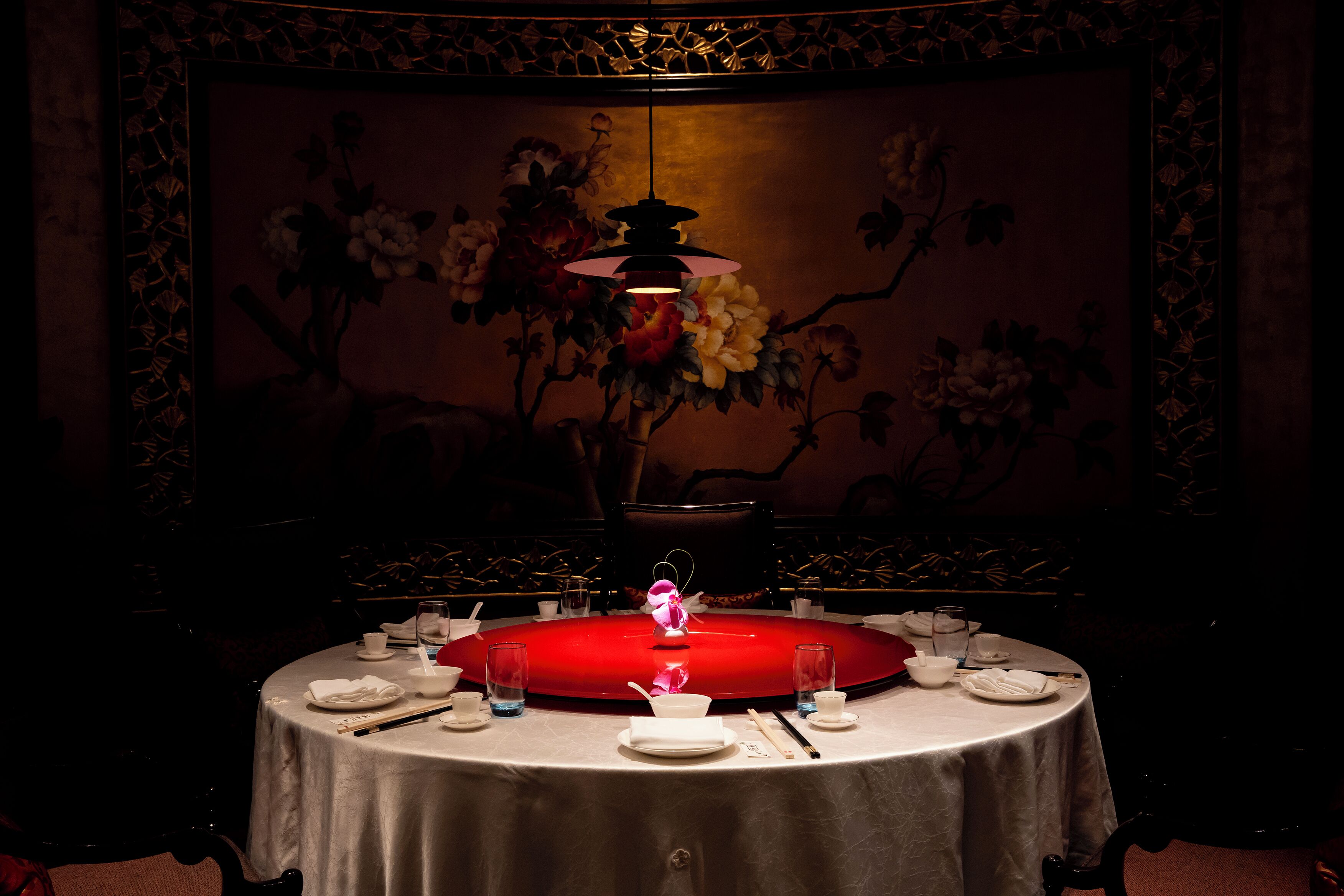 Mesa de jantar posta para uma refeição, com um centro de mesa vermelho, rodeada por cadeiras e um quadro tradicional na parede.