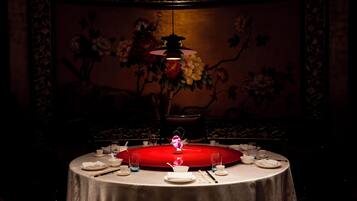 Mesa de jantar posta para uma refeição, com um centro de mesa vermelho, rodeada por cadeiras e um quadro tradicional na parede.