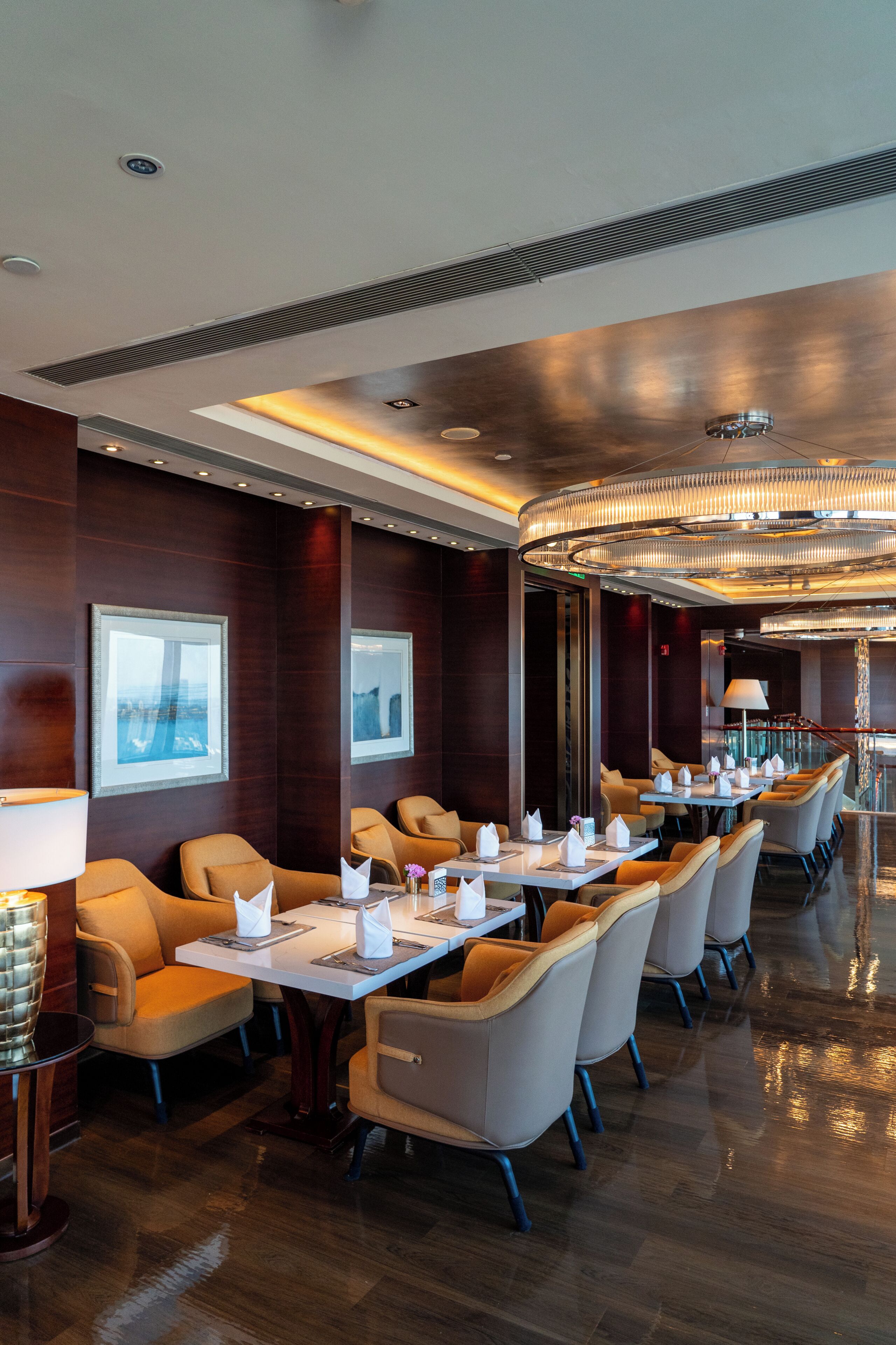 Uma sala de jantar de hotel com mesas compridas, cadeiras e um lustre moderno.