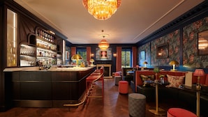 Uma área de bar com balcão de madeira, banquetas e um lustre grande. Há várias mesas e cadeiras dispostas para acomodar os clientes.