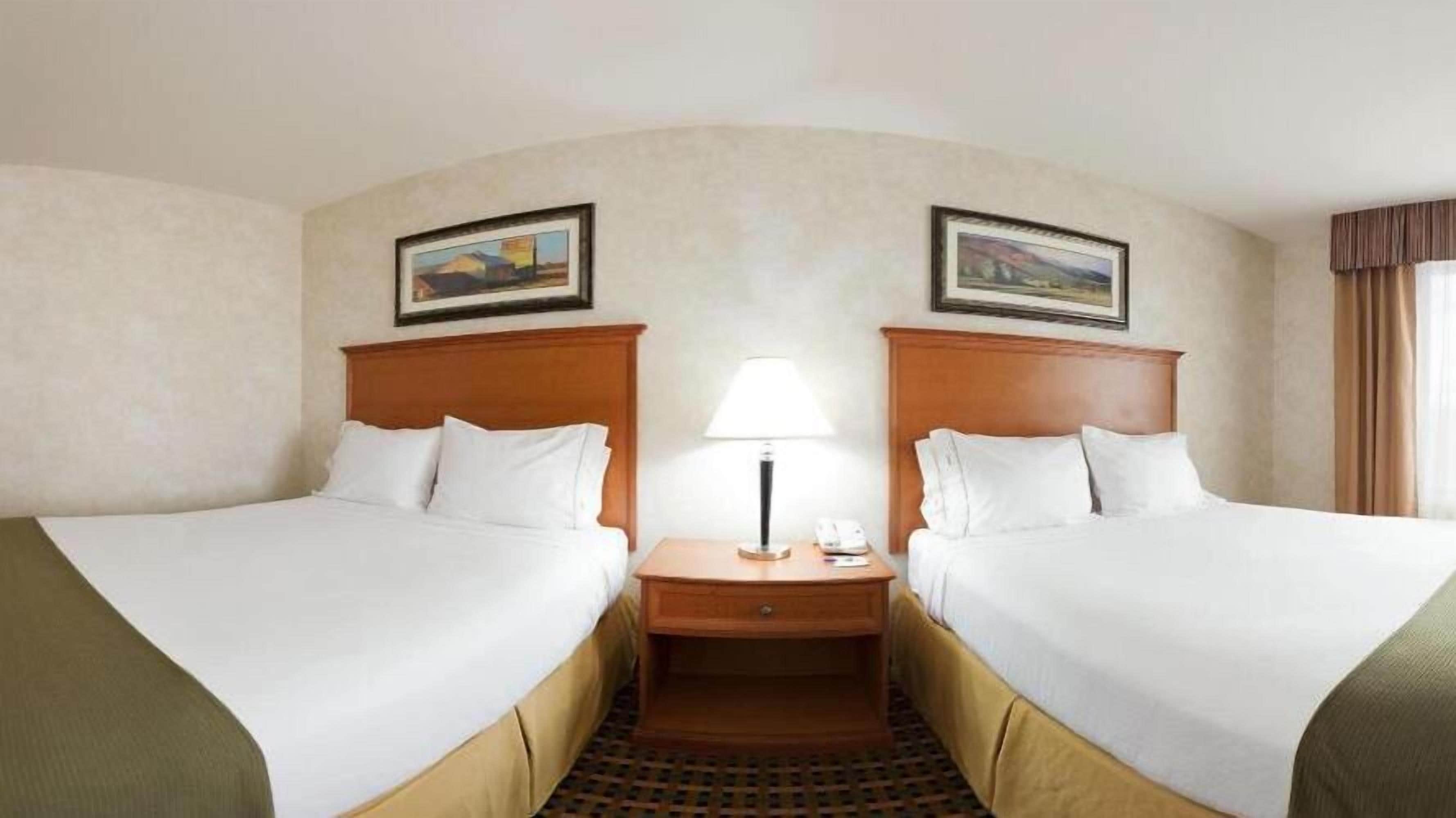 Quarto de hotel com duas camas, uma mesa de cabeceira com abajur e duas fotos emolduradas na parede.