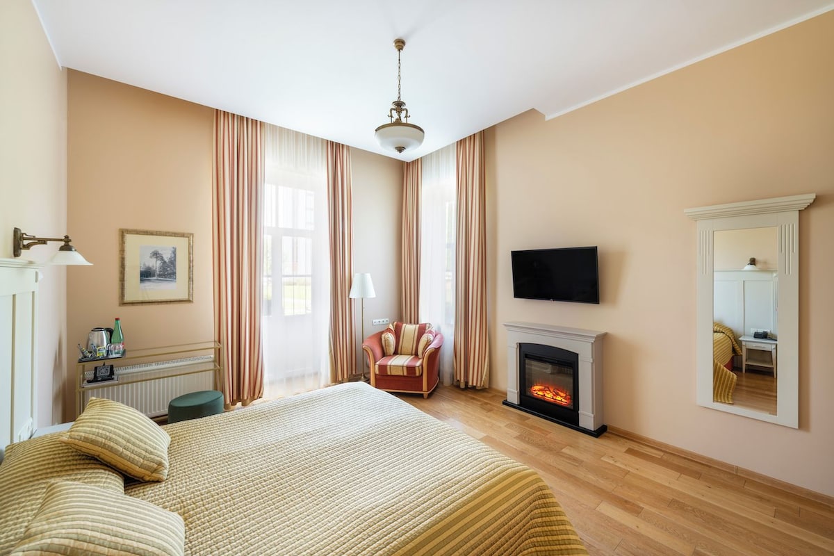 Quarto de hotel com cama, lareira, televisão e uma grande janela com cortinas.