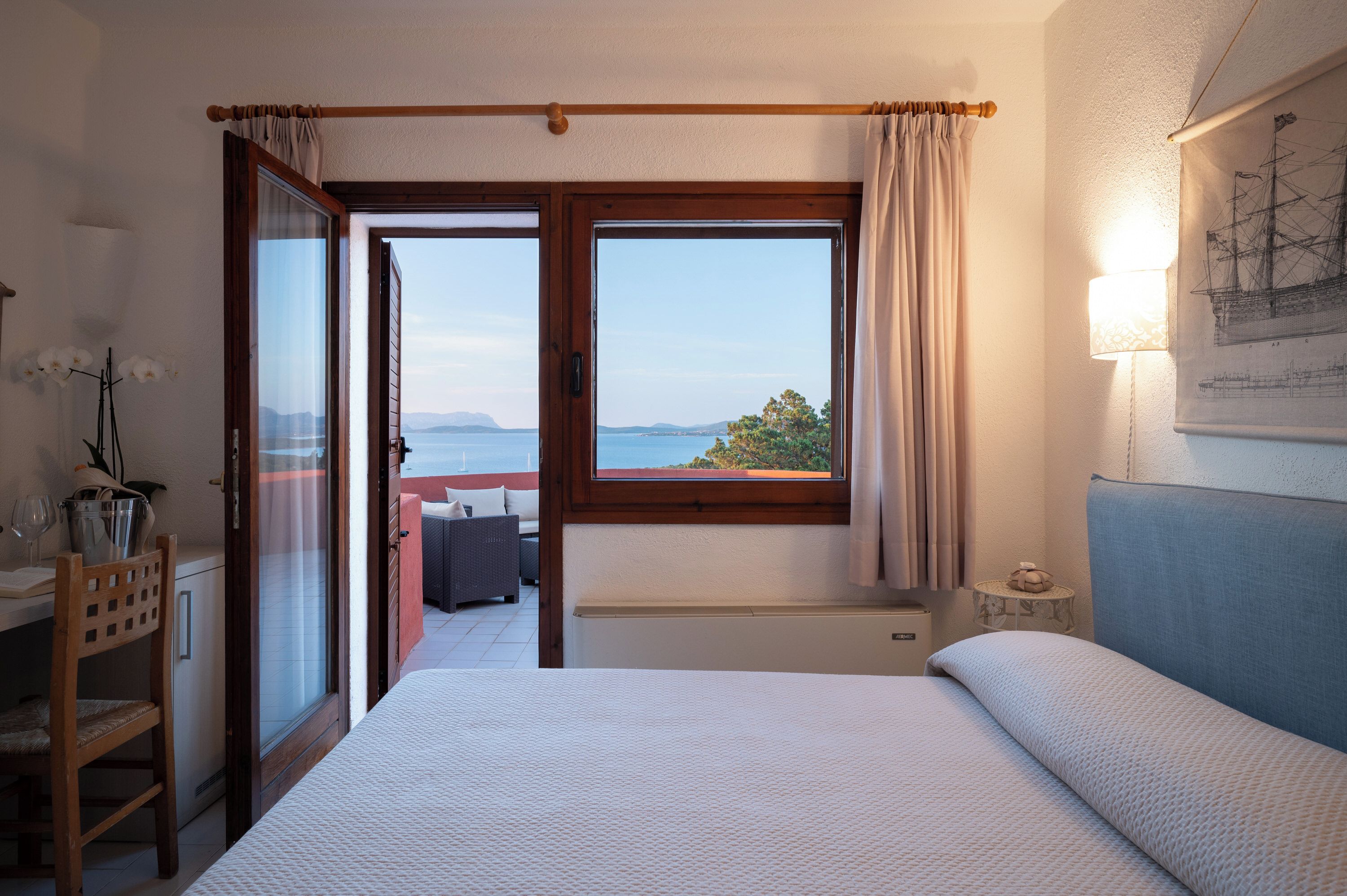 Quarto de hotel com cama, escrivaninha com cadeira, janela com cortinas e vista para o mar.