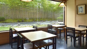 Um restaurante com mesas e cadeiras de madeira, grandes janelas com vista para uma floresta de bambu.