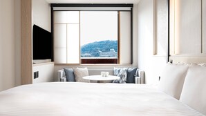 Quarto de hotel moderno com uma cama grande, uma mesa redonda pequena e uma janela com vista para a paisagem urbana e colinas.