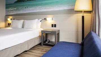 Quarto de hotel com cama, mesa de cabeceira, abajur e uma pintura de uma cena de praia.