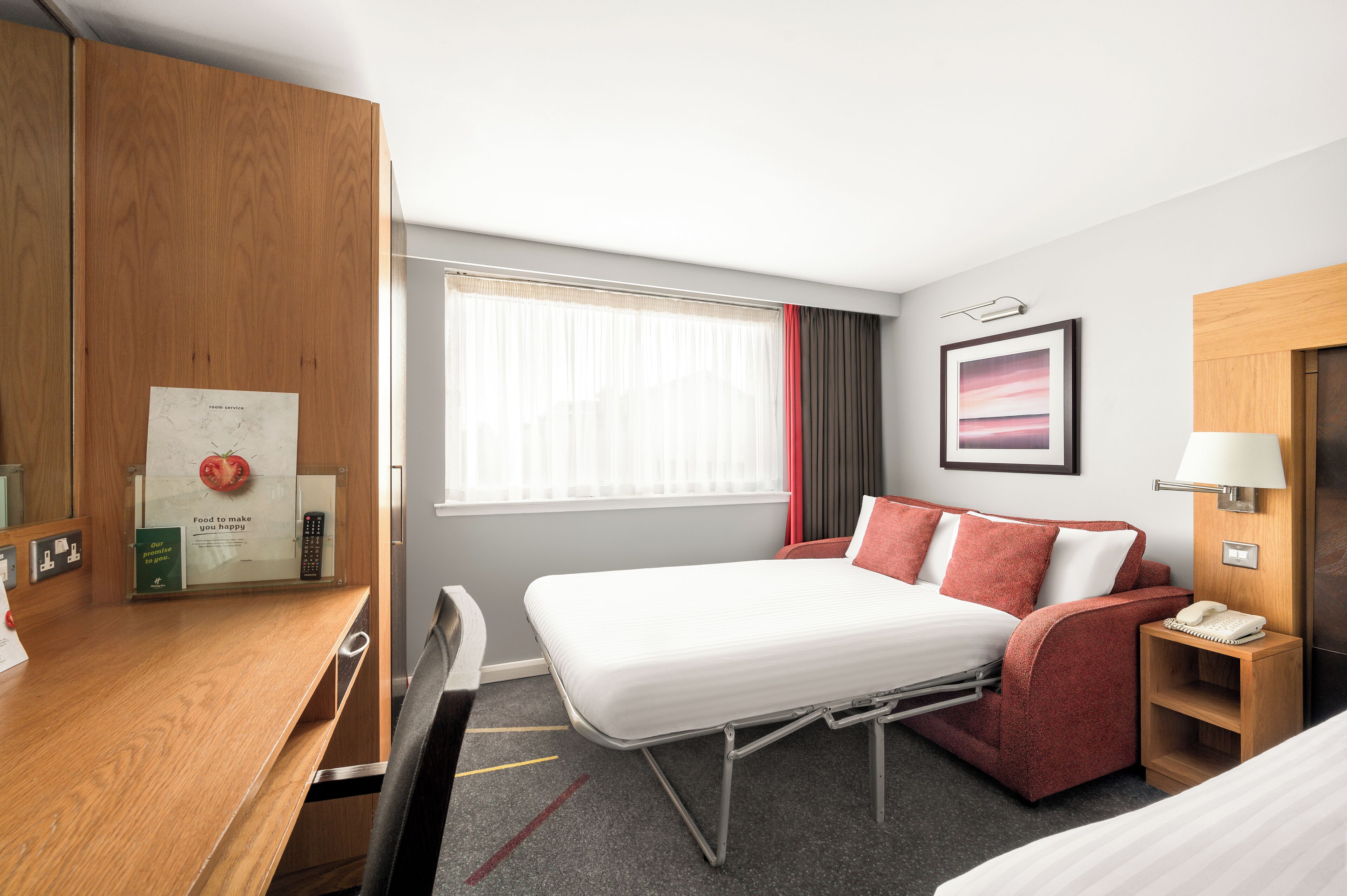 Quarto de hotel com cama, escrivaninha, cadeira, mesa de cabeceira com abajur, quadro na parede e janela com cortinas.