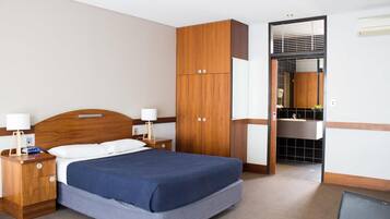 Quarto de hotel com cama, guarda-roupa de madeira, mesas de cabeceira e banheiro com pia e espelho.