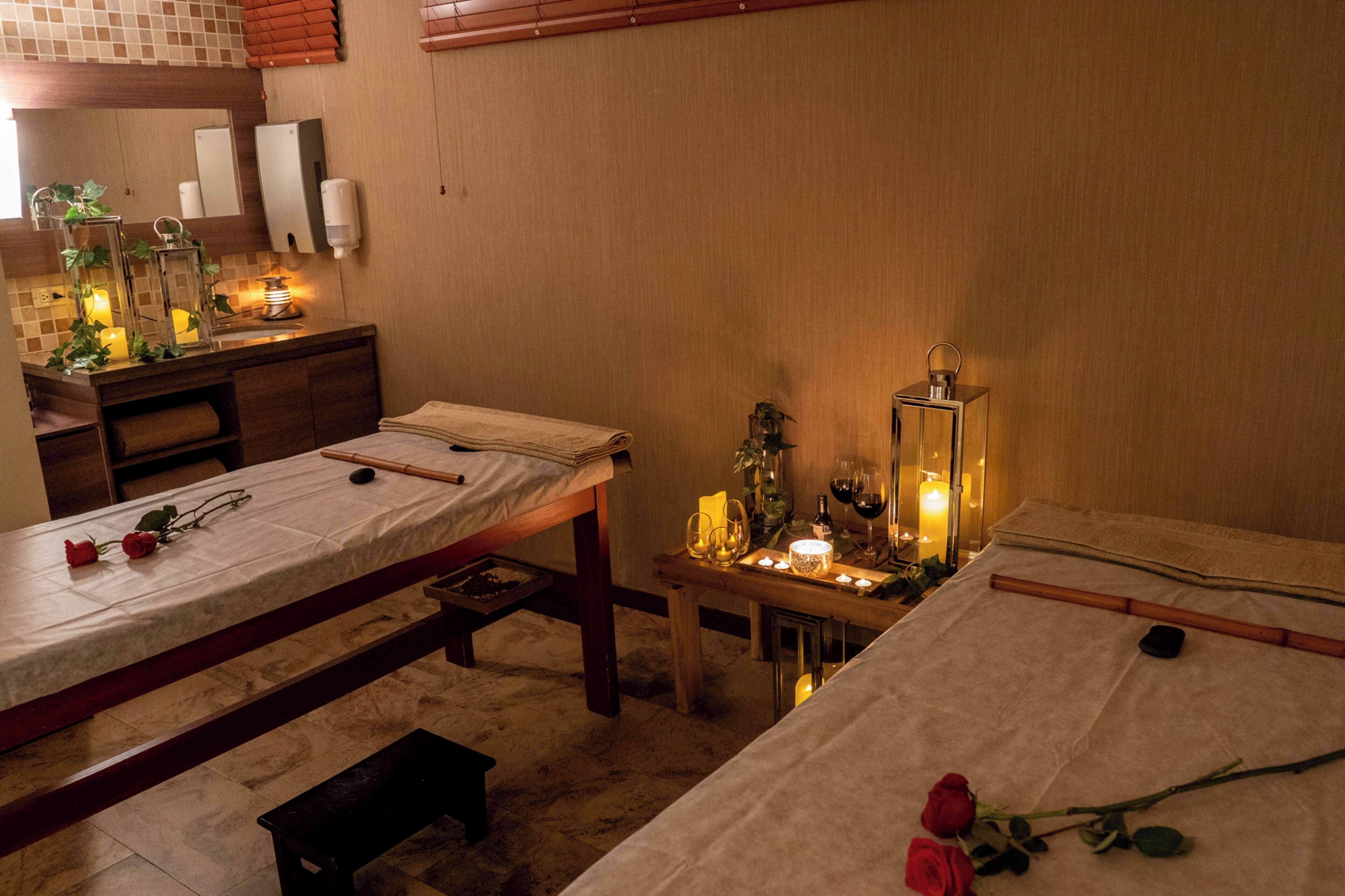Sala de spa com duas macas de massagem, velas e uma pequena mesa com garrafa e copo.