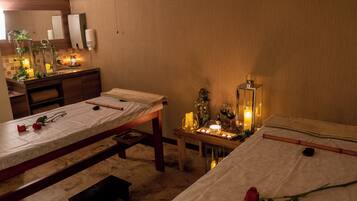 Sala de spa com duas macas de massagem, velas e uma pequena mesa com garrafa e copo.