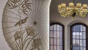 Quarto com um grande mural na parede retratando bambu e um pássaro, um candelabro e duas janelas em arco com uma parede de tijolos visível ao fundo.