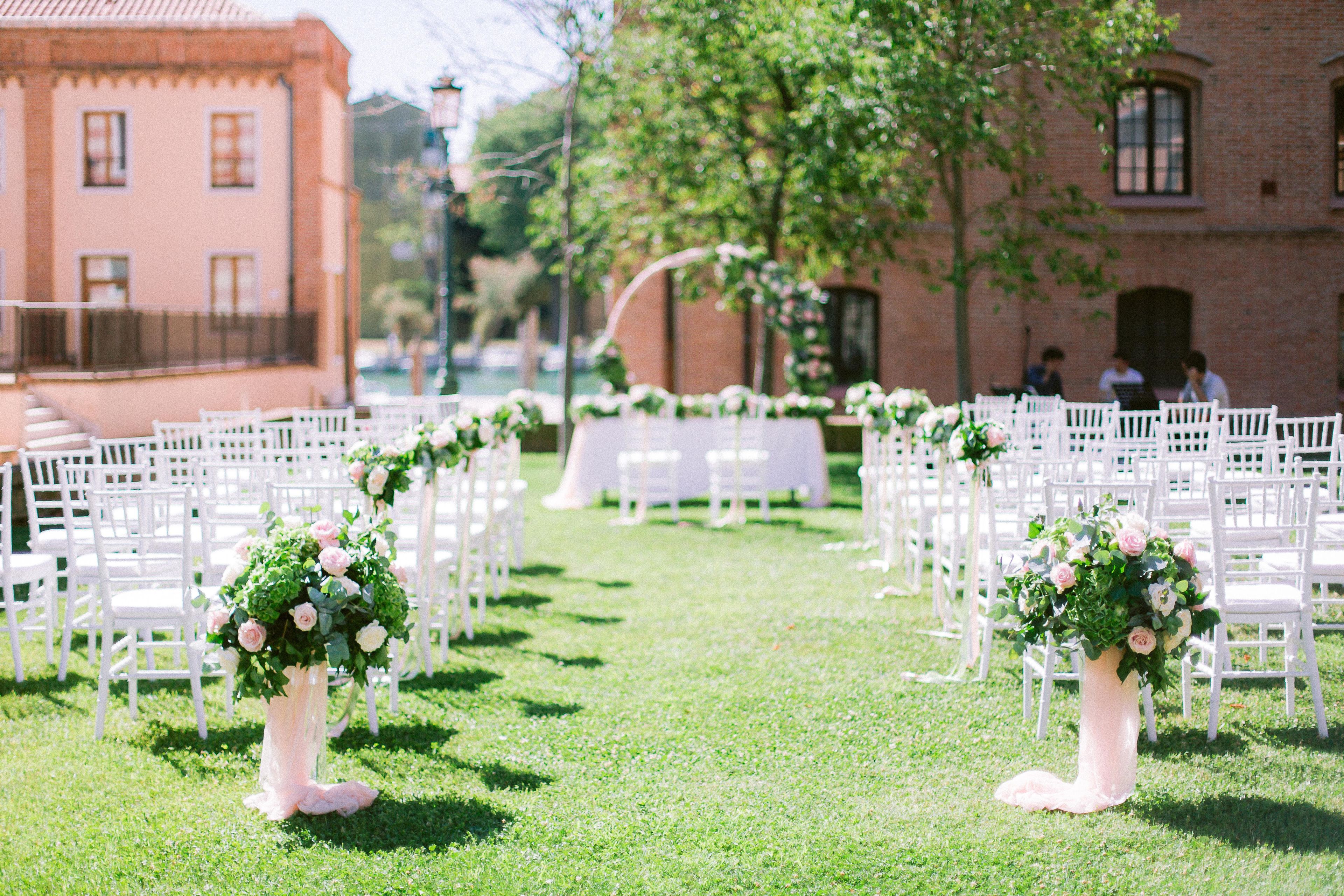 Espaço para casamento ao ar livre, com cadeiras brancas dispostas em fileiras, área gramada e um edifício ao fundo.