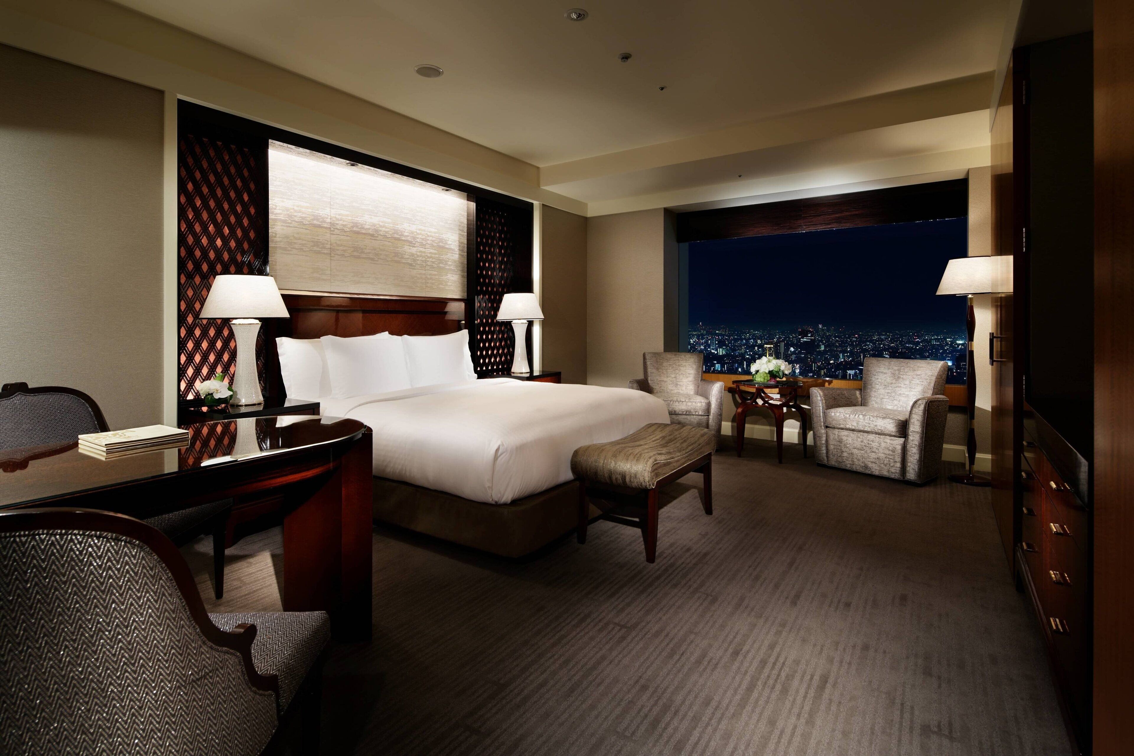 Quarto de hotel com uma cama grande, duas poltronas, uma escrivaninha e vista para a cidade à noite.