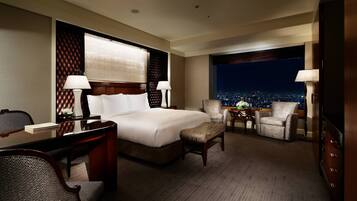 Quarto de hotel com uma cama grande, duas poltronas, uma escrivaninha e vista para a cidade à noite.