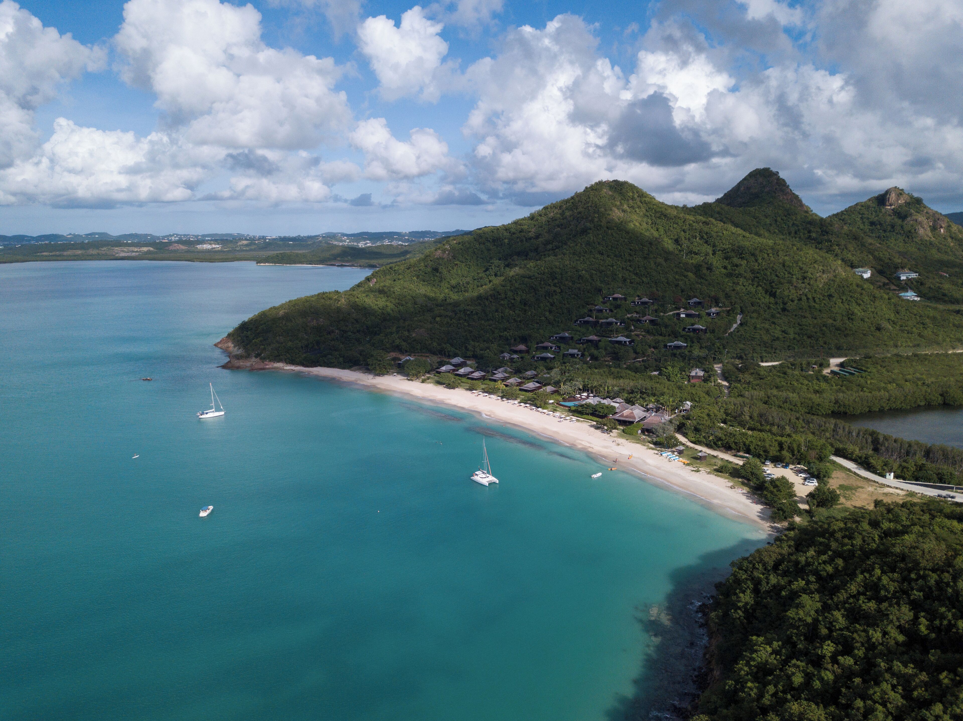 Vista aérea de uma área costeira com praia, águas turquesa cristalinas e um pequeno povoado com casas numa encosta.