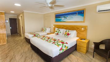 Quarto de hotel com duas camas, ventoinha no teto, ar condicionado, cabeceira de madeira, mesa de cabeceira, uma cadeira e uma pintura de uma paisagem praiana.