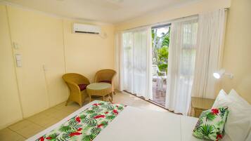 Quarto de hotel com cama, duas cadeiras de vime, uma mesinha e vista para o exterior.