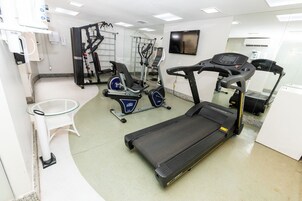 Academia com máquinas de cardio, equipamentos de musculação e uma TV na parede.