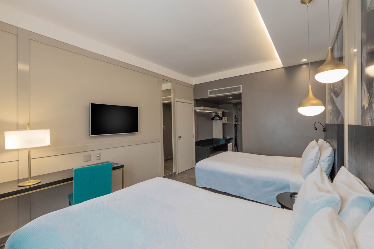 Quarto de hotel com duas camas, uma escrivaninha, televisão e banheiro visível através de uma porta aberta.
