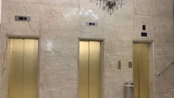 Um hall de entrada com piso de mármore, três elevadores dourados e uma lixeira.