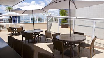 Pátio externo com mesas redondas e cadeiras de vime, guarda-sóis e vista para uma piscina e edifícios.