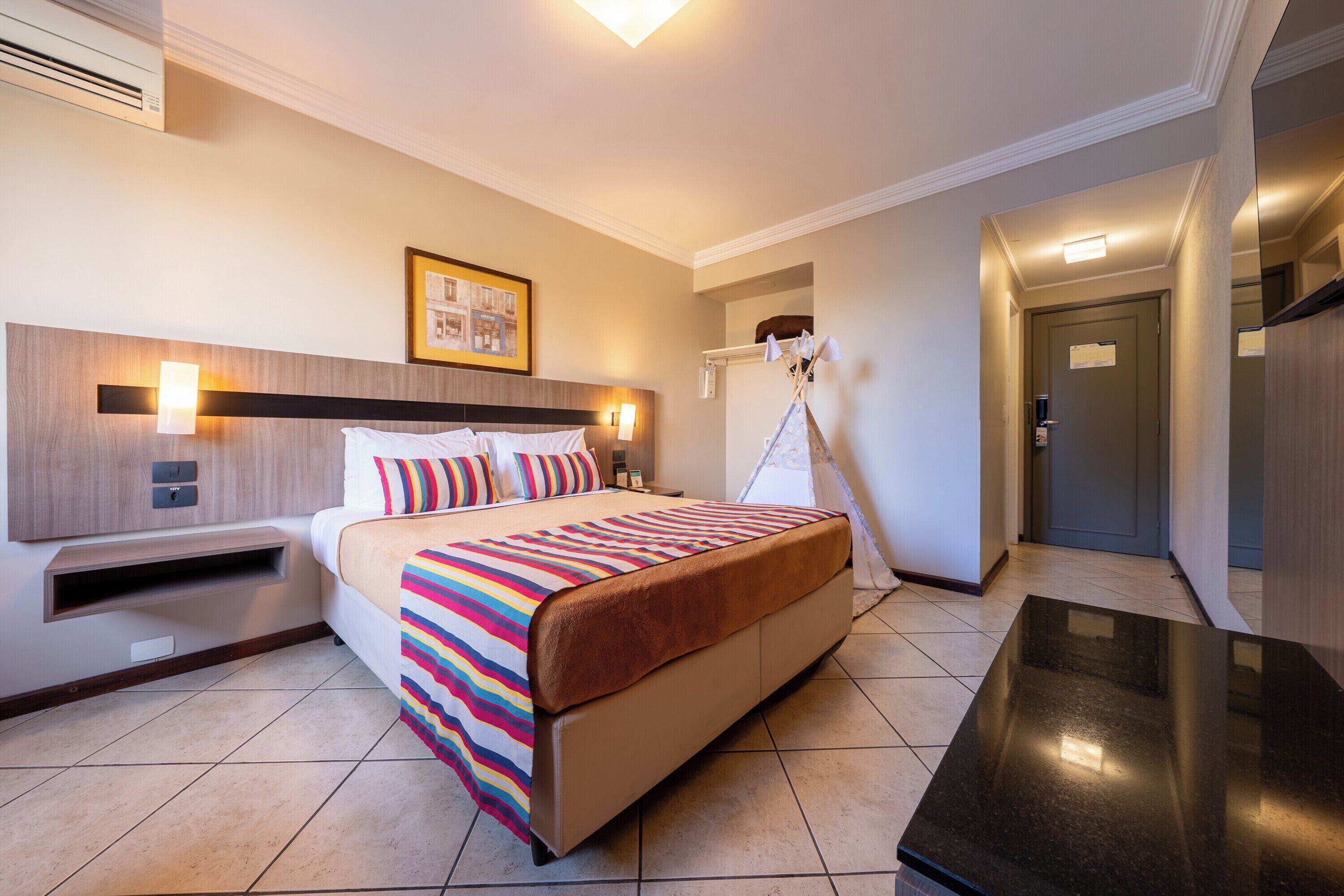 Quarto de hotel com cama, mesas de cabeceira, cabeceira, ar condicionado fixado na parede, quadro emoldurado, janela com vista e uma porta.