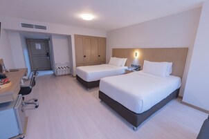 Quarto de hotel com duas camas, uma escrivaninha com cadeira, um guarda-roupa e uma mesa de cabeceira com abajur.