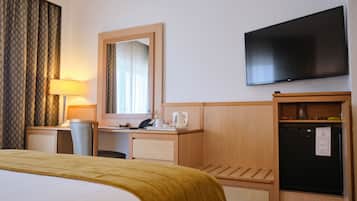 Quarto de hotel com cama, escrivaninha, cadeira, luminária, espelho, televisão e frigorífico.