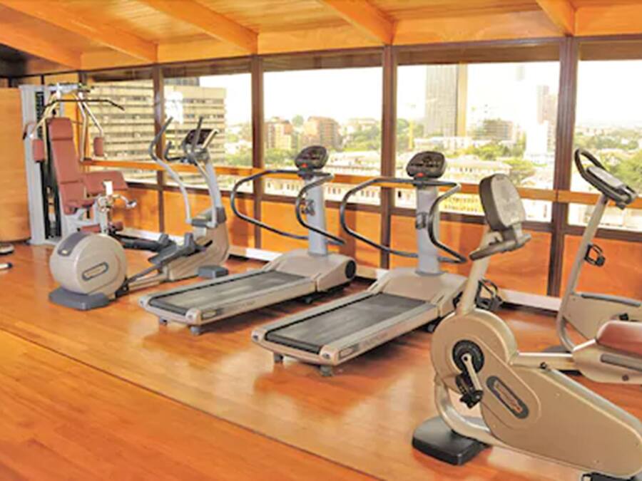 Academia com diversos equipamentos de exercício, grandes janelas e piso de madeira.