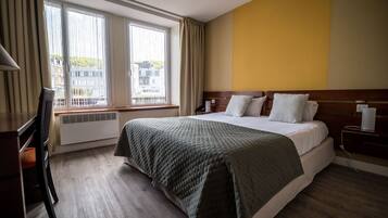 Quarto de hotel com cama, escrivaninha e cadeira. Há uma janela com cortinas e vista para edifícios.