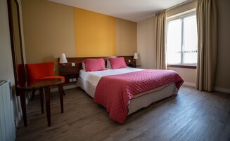 Quarto de hotel com cama, cadeira, mesa de cabeceira e janela com cortinas.