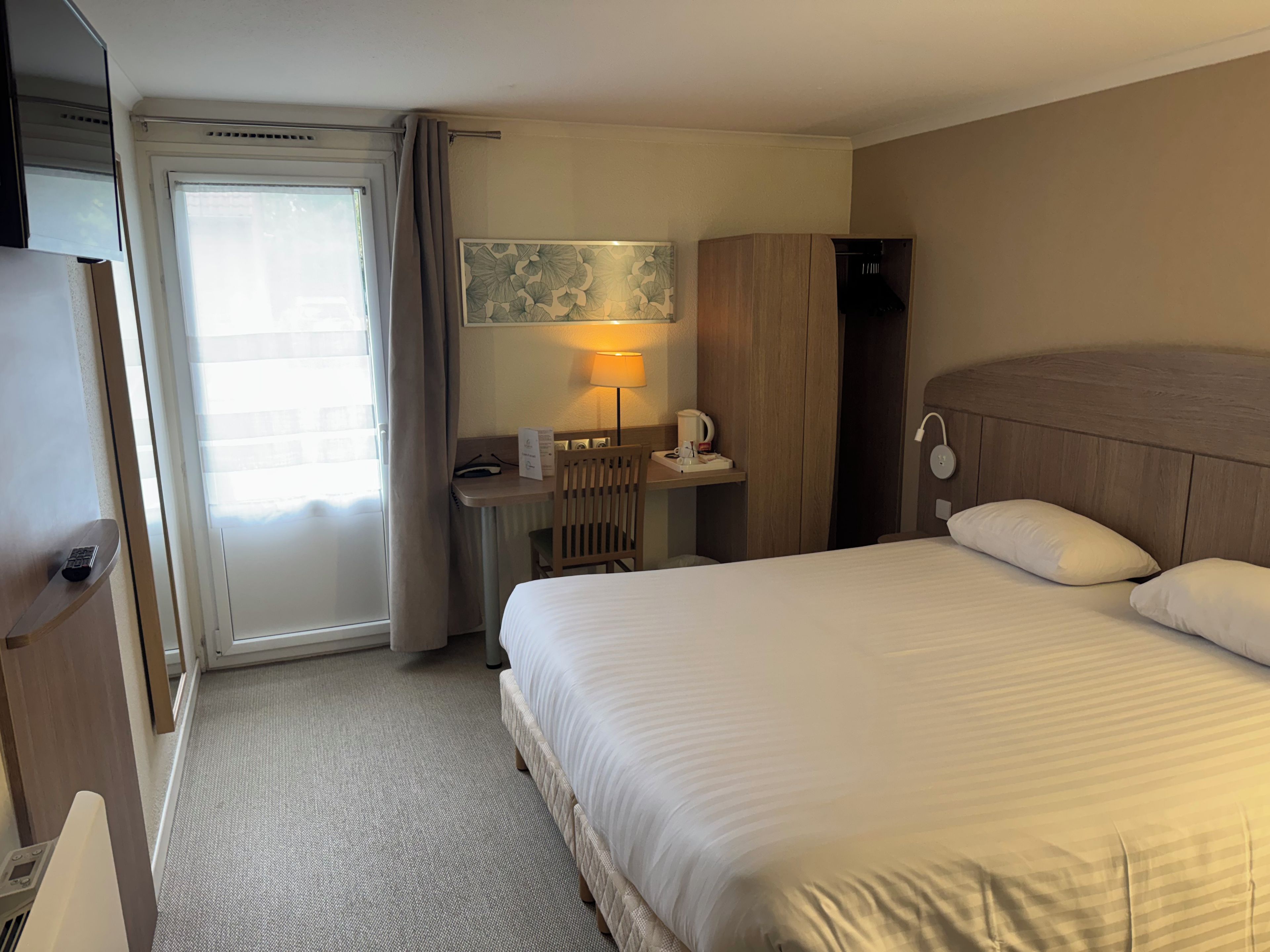 Quarto de hotel com cama, escrivaninha com luminária, guarda-roupa e porta que dá para uma varanda.