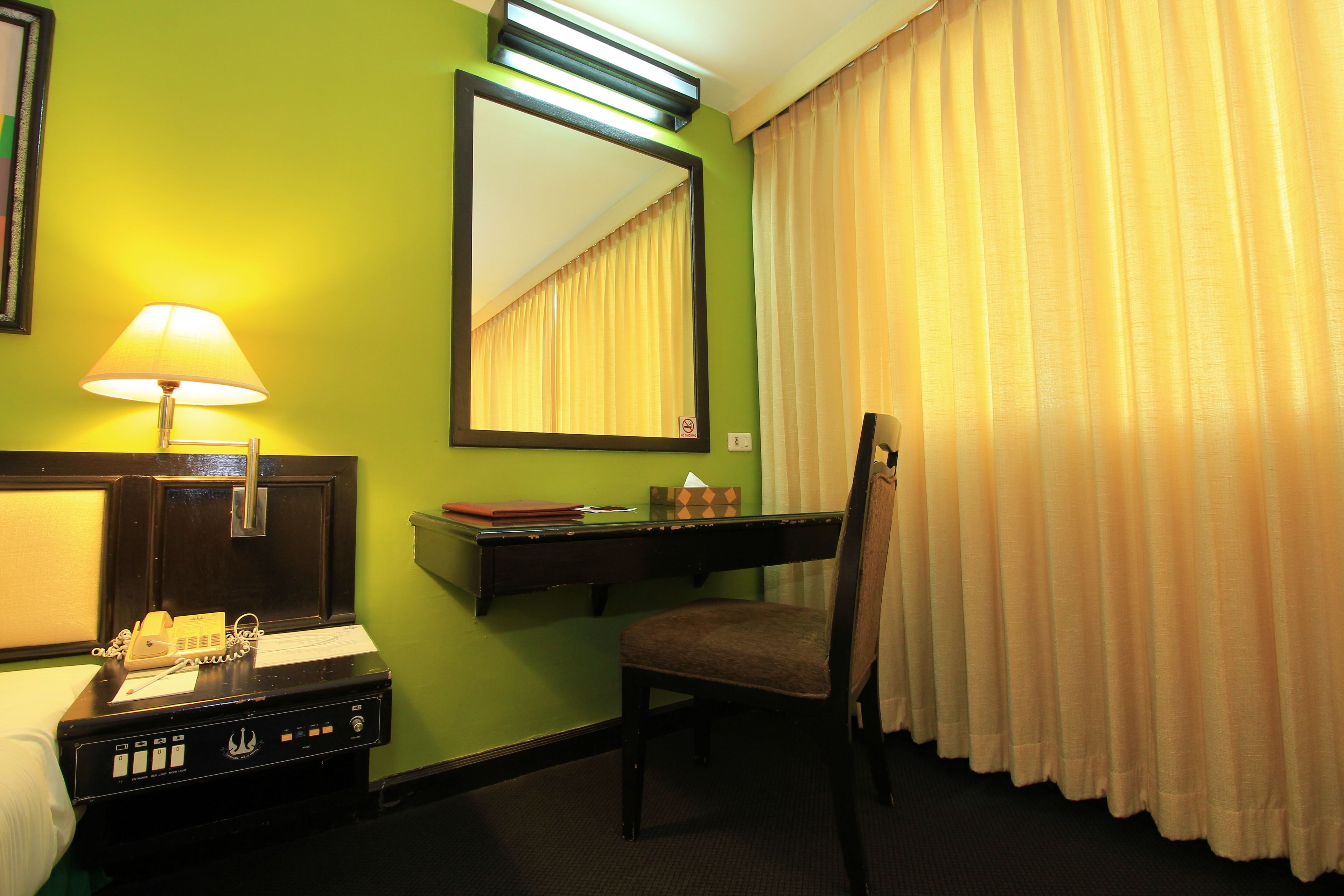 Quarto de hotel com cama, escrivaninha com luminária, cadeira, espelho e janela com cortinas.