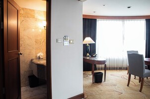 Quarto de hotel com banheiro de mármore, área de jantar com mesa e cadeiras e uma grande janela com cortinas.