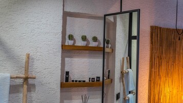 Um banheiro com parede branca texturizada, prateleiras de madeira com artigos de higiene pessoal, um espelho e um painel de parede de bambu.