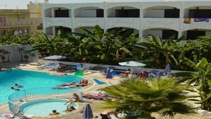 Uma área de piscina no terraço com várias espreguiçadeiras e guarda-sóis, cercada por vegetação exuberante e um edifício alto ao fundo.