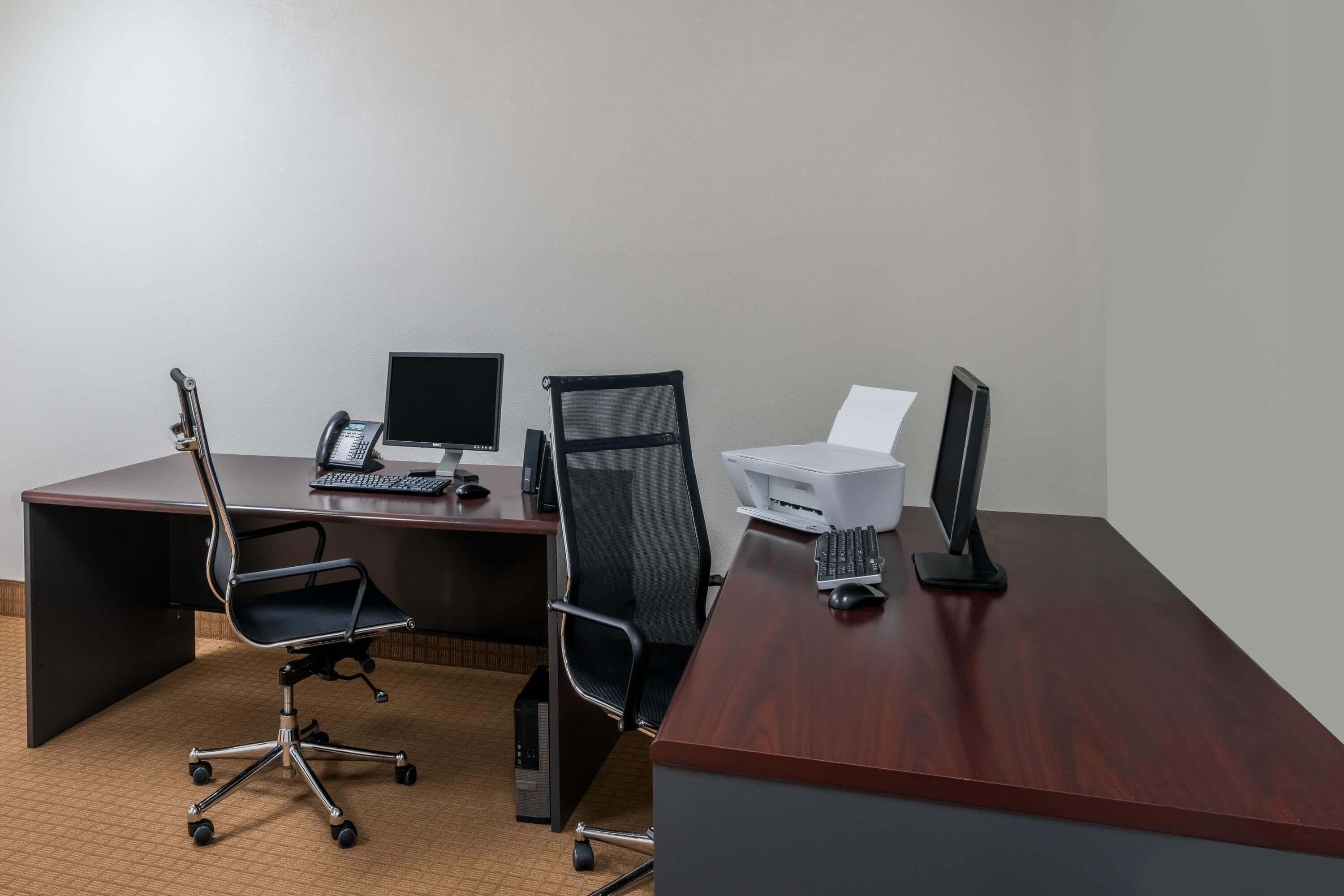 Sala de conferência com uma grande mesa de madeira, duas cadeiras de escritório pretas, um computador de mesa, um monitor, uma impressora e um telefone.