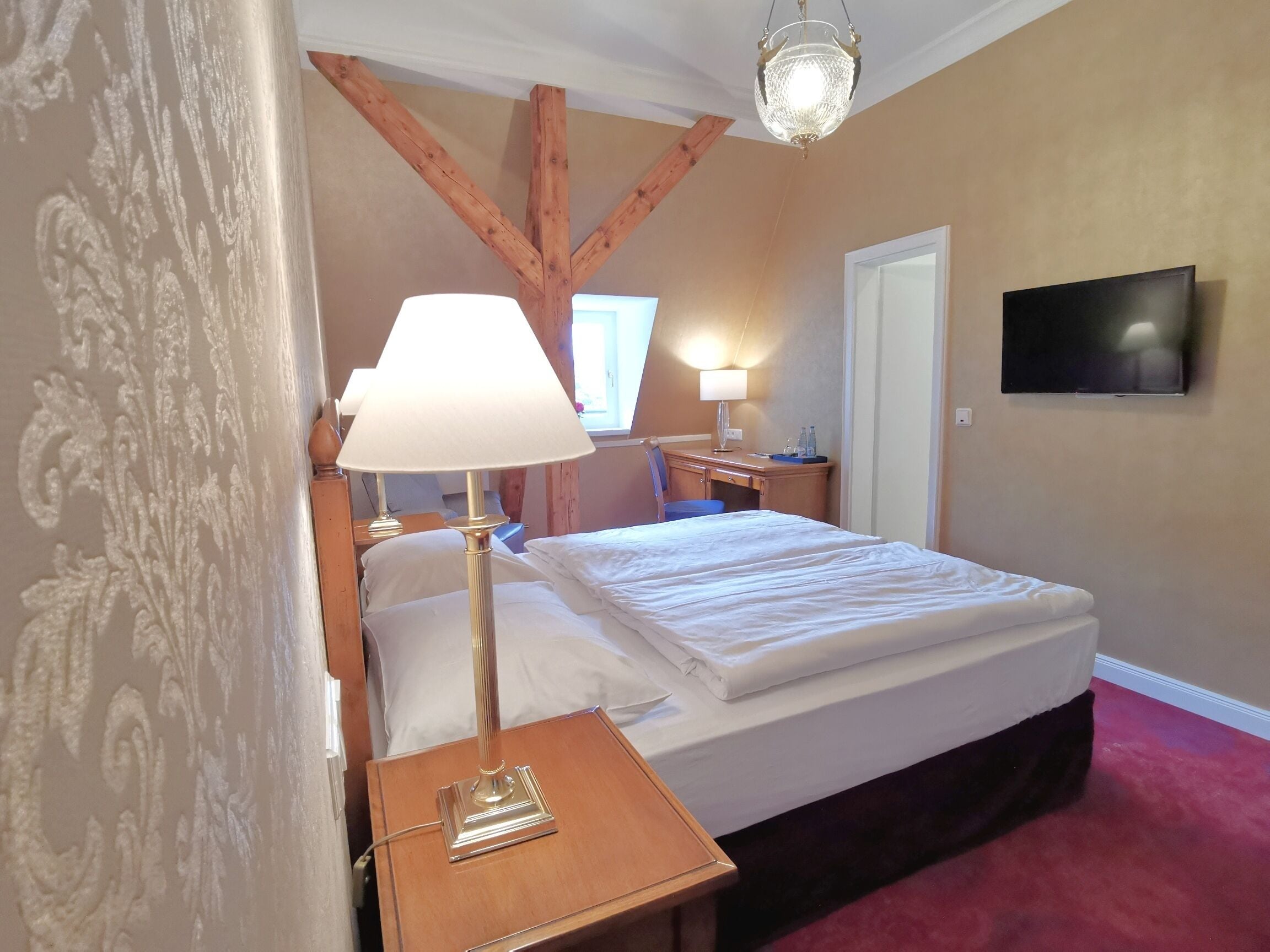Quarto de hotel com cama, mesas de cabeceira, escrivaninha, cadeira, televisão e lustre.