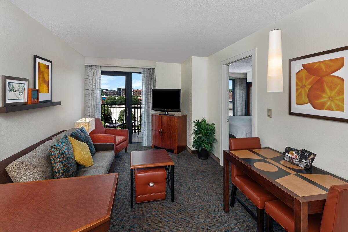 Quarto de hotel moderno com sofá, mesa de jantar com bancos, televisão de tela plana e varanda com vista para a cidade.