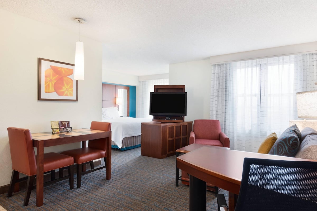 Quarto de hotel com uma cama, televisão, área de refeições com cadeiras, sofá e uma grande janela com cortinas.